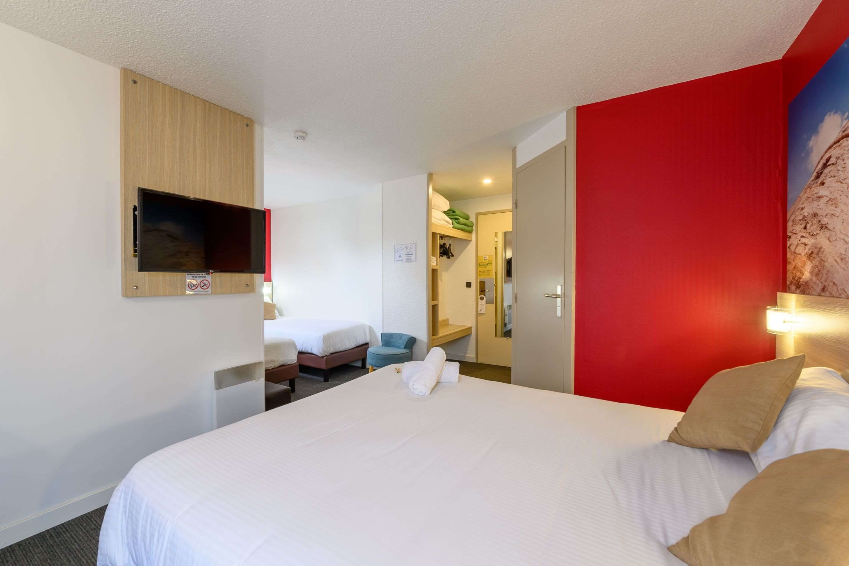 Quarto de hotel com cama, televisão presa na parede, escrivaninha com cadeira e parede em destaque vermelha.