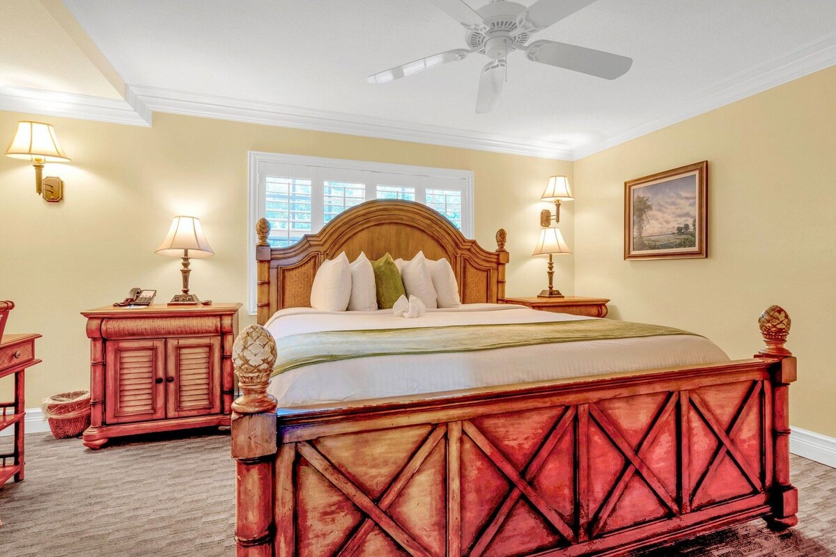 Quarto com cama de madeira, duas mesas de cabeceira com luminárias, ventilador de teto e janela com vista.
