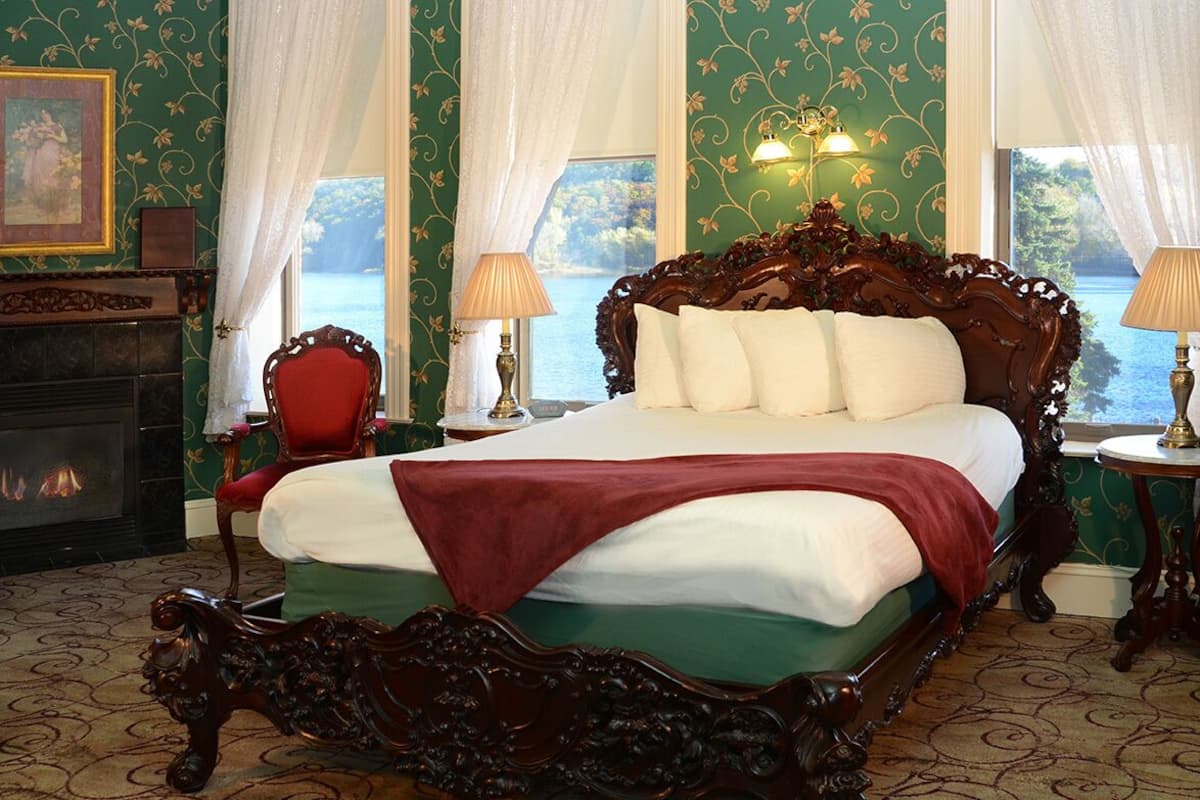 Uma cama grande e ornamentada com colcha vermelha e branca em um quarto com papel de parede floral verde, lareira e vista para um corpo d'água.