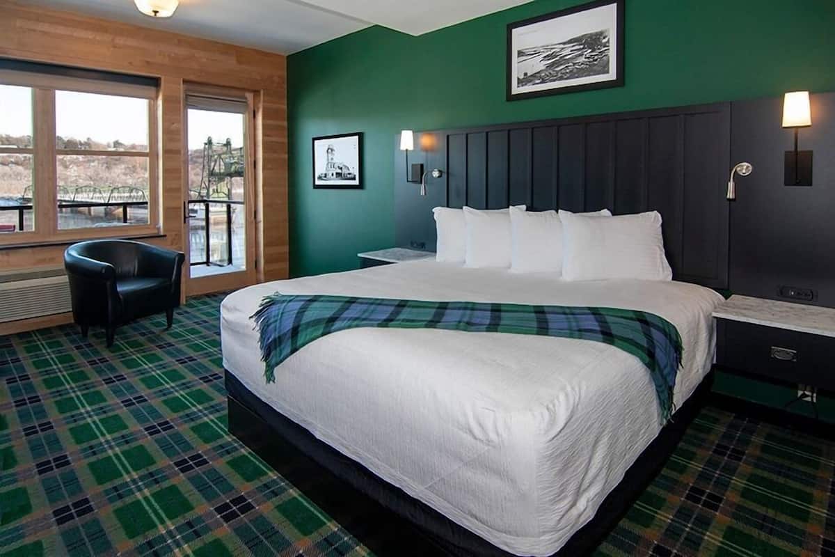 Quarto de hotel com uma cama grande, uma cadeira, uma mesinha de cabeceira e vista para o exterior pela janela.