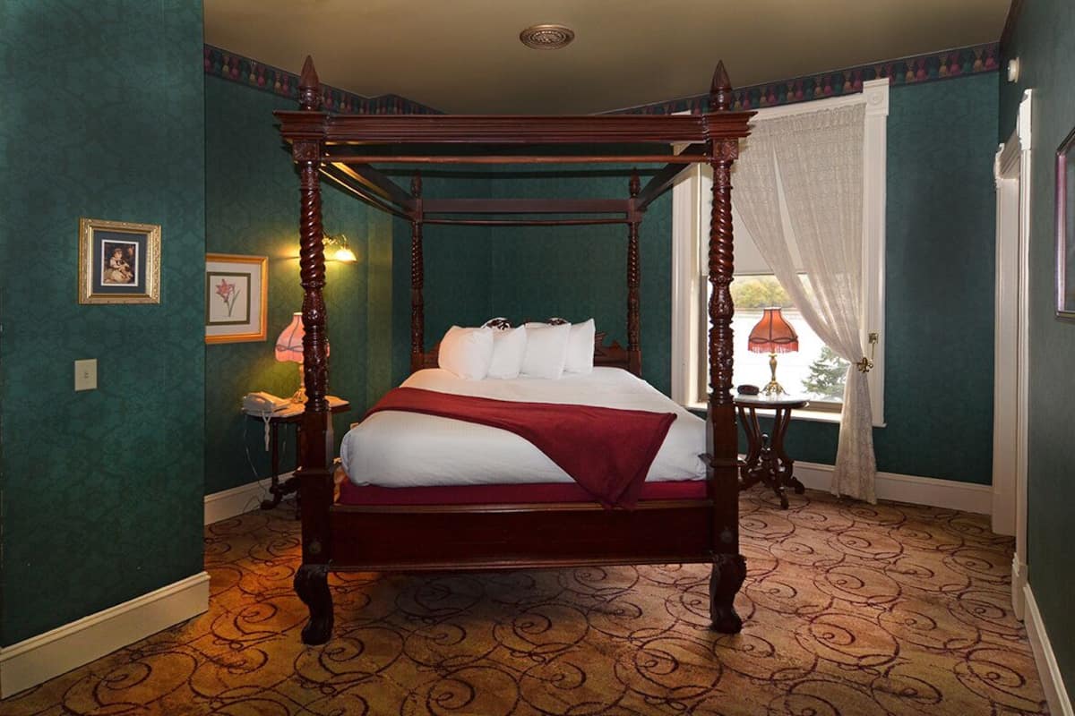 Uma cama com dossel em um quarto com paredes verde-água, carpete estampado e uma janela com cortinas brancas.