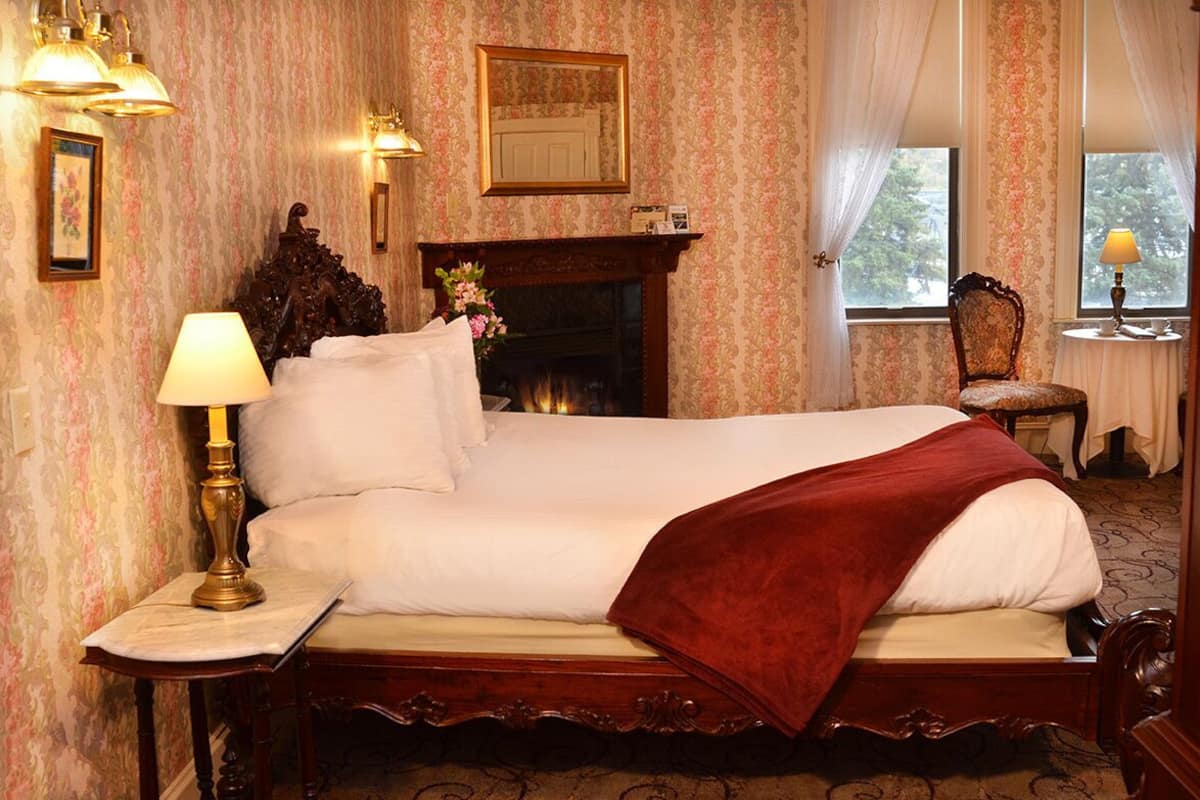 Quarto de hotel clássico com uma cama grande, lareira, mesa de cabeceira com abajur e uma cadeira perto da janela.