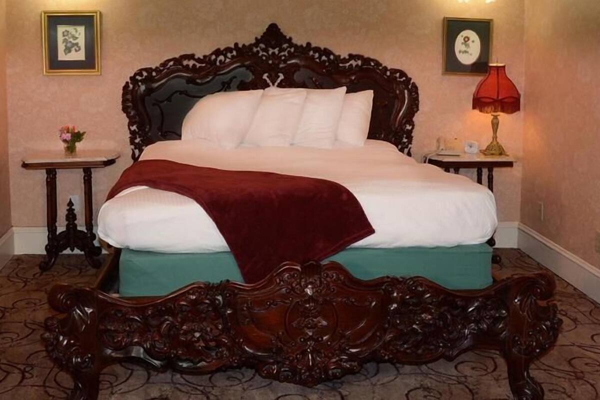 Uma cama grande e elaborada, com estrutura de madeira escura e colchão branco, acompanhada por duas mesinhas de cabeceira com luminárias.