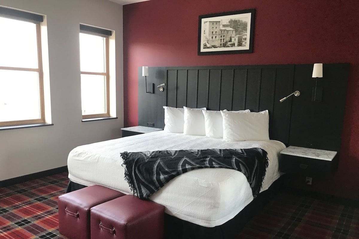 Quarto de hotel com uma cama grande, cabeceira, mesa de cabeceira, janela, tapete xadrez vermelho e mala vermelha.
