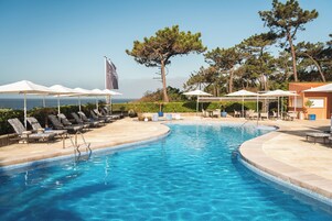 Área de piscina com espreguiçadeiras e guarda-sóis, cercada por árvores e vista desobstruída do oceano.