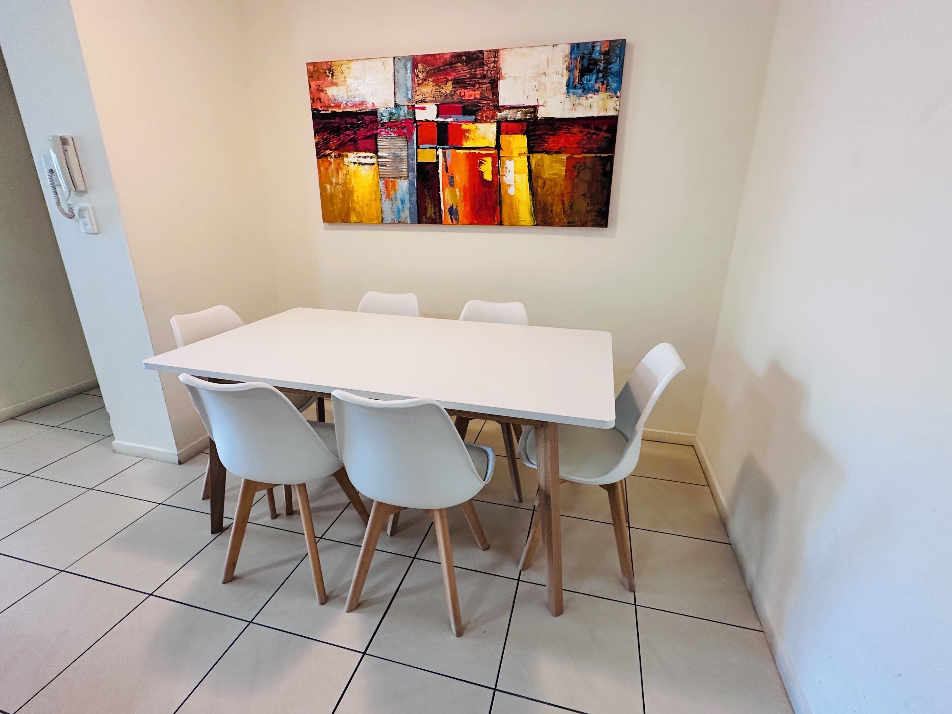 Uma sala de jantar com uma mesa branca e quatro cadeiras, um quadro abstrato colorido na parede e um chão ladrilhado.