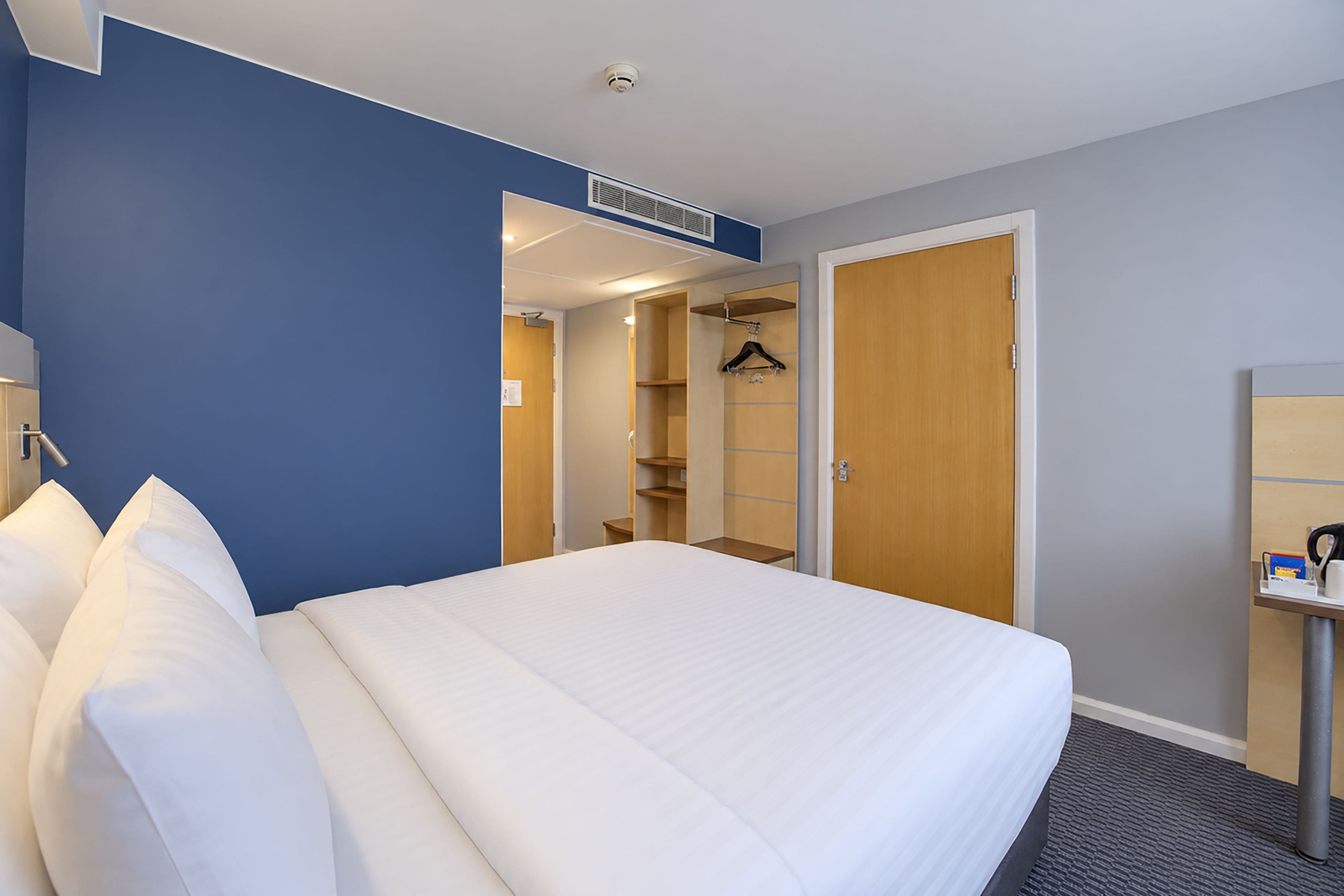Quarto de hotel com uma cama grande, guarda-roupa, espelho e secretária com chaleira.