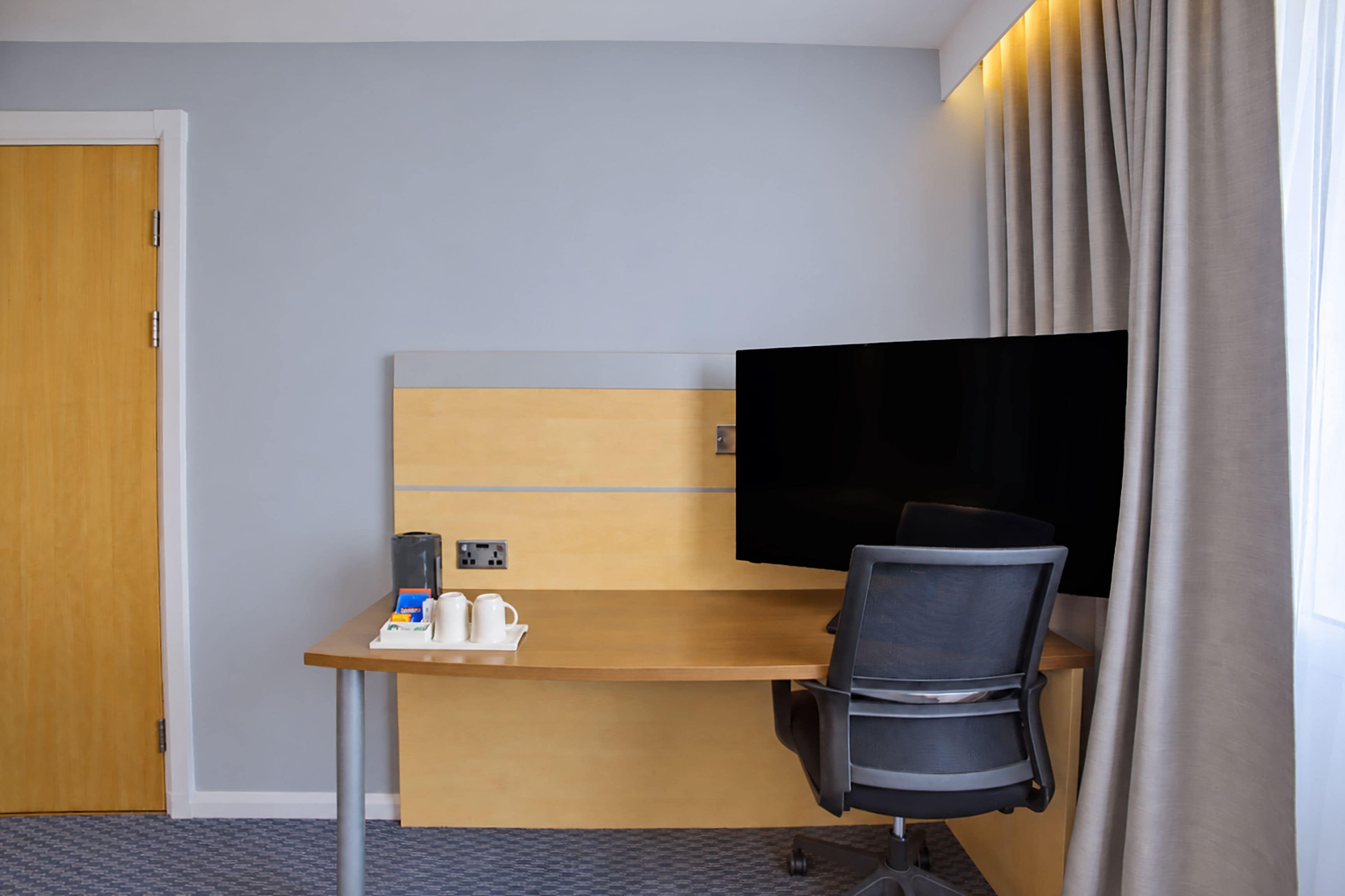 Quarto de hotel com escrivaninha, uma cadeira, televisão e porta.