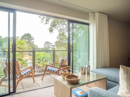 Quarto com mesa de vidro, cadeiras de madeira, um sofá e vista para o verde lá fora.