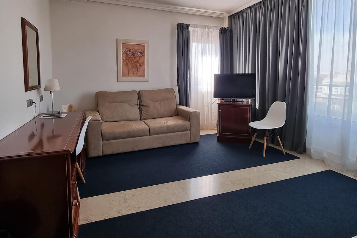 Quarto de hotel com sofá, televisão sobre suporte de madeira, cadeira branca, escrivaninha com luminária e pintura na parede.