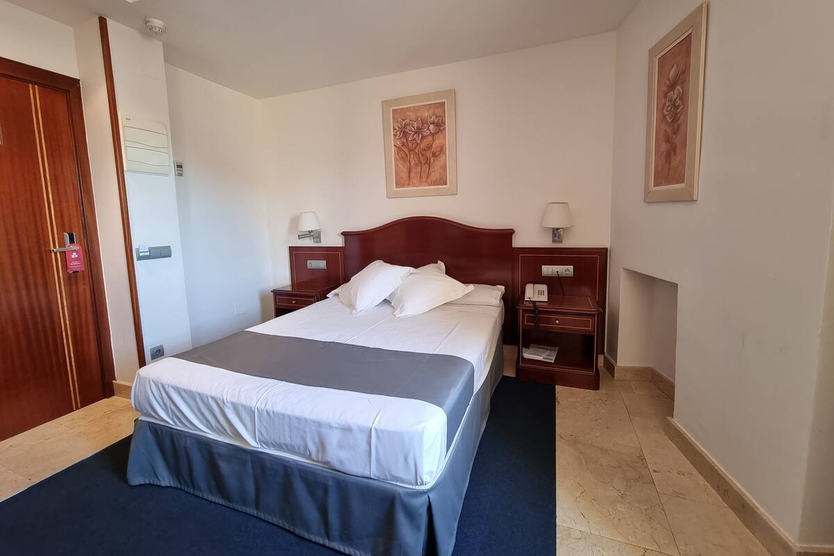 Quarto de hotel com cama grande, cabeceira de madeira, mesas de cabeceira com luminárias e obras de arte nas paredes.