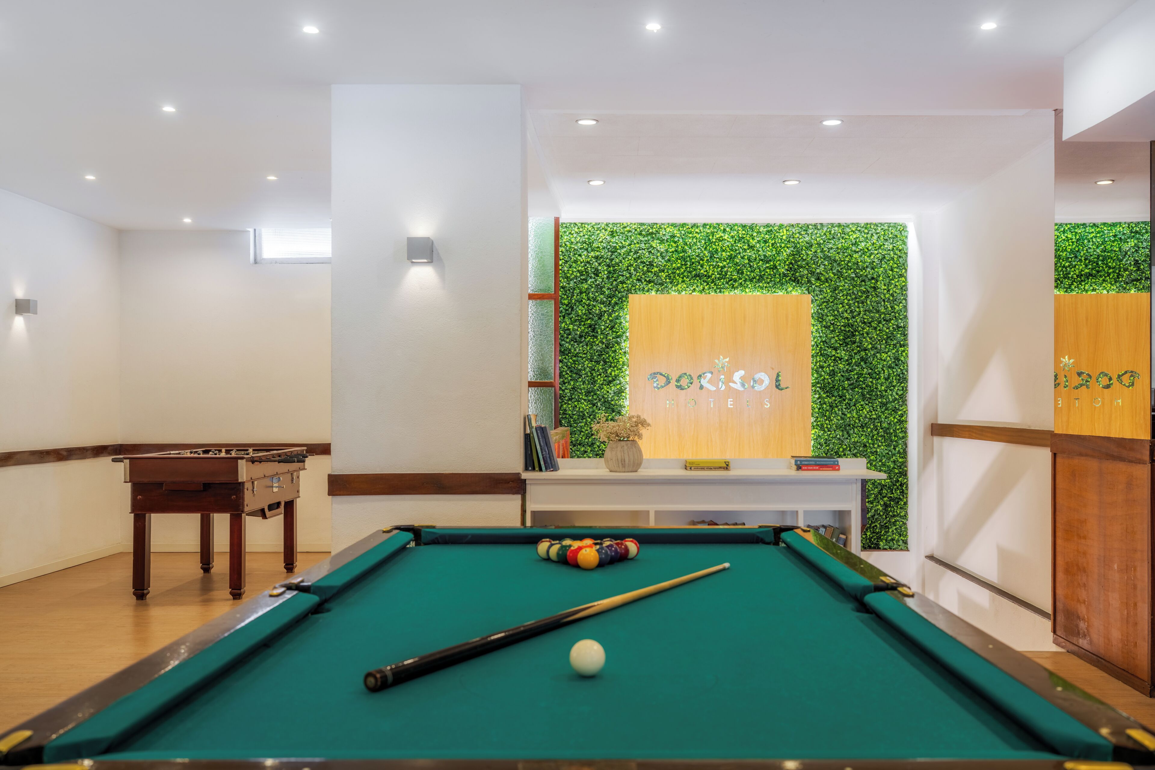 Mesa de bilhar com bolas e taco, mesa de futebol de mesa, balcão de recepção com uma placa e parede verde com painel de madeira.