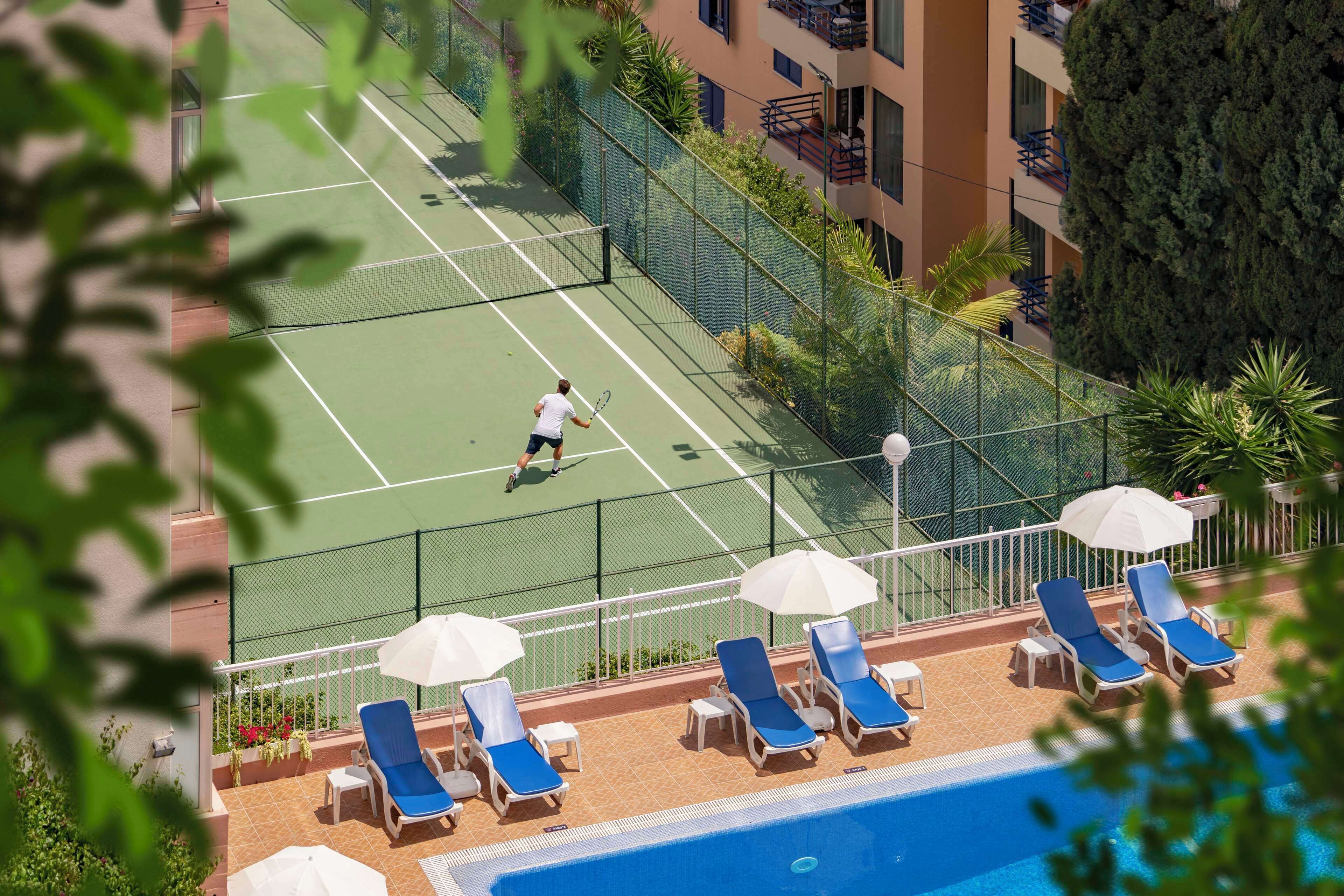 Uma quadra de tênis com rede, cercada por área de piscina com espreguiçadeiras e guarda-sóis.