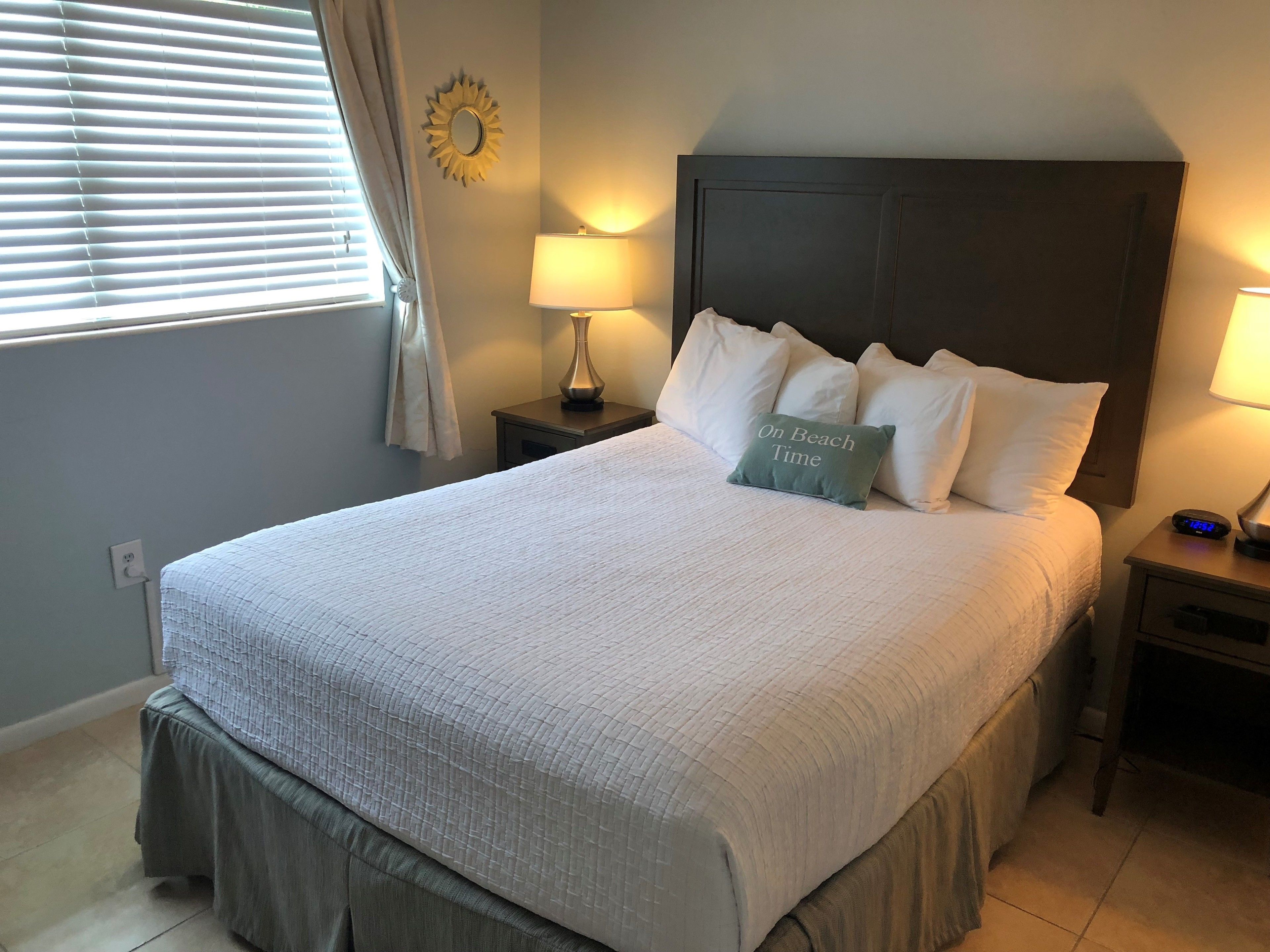 Cama bem arrumada com roupa de cama branca e um travesseiro verde com texto, uma luminária de cabeceira, mesa de cabeceira com relógio e uma janela com persianas.