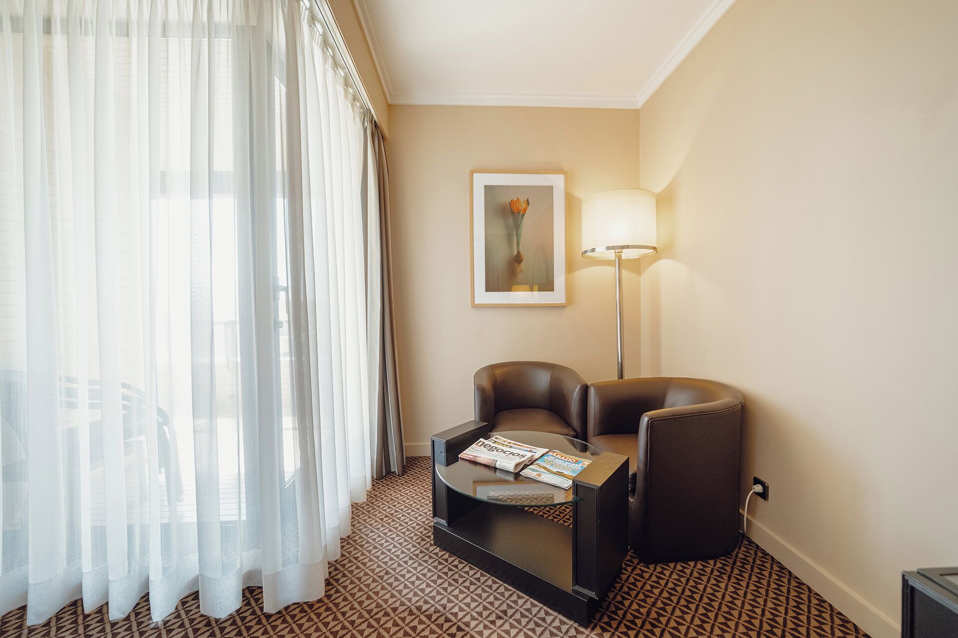Quarto de hotel com mesa de centro de vidro, duas poltronas de couro, abajur e quadro na parede.