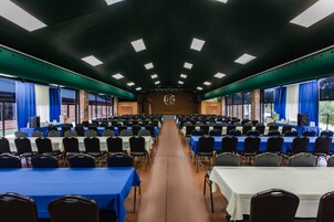Uma sala de conferências com fileiras de mesas e cadeiras, um palco na frente e janelas amplas em ambos os lados.