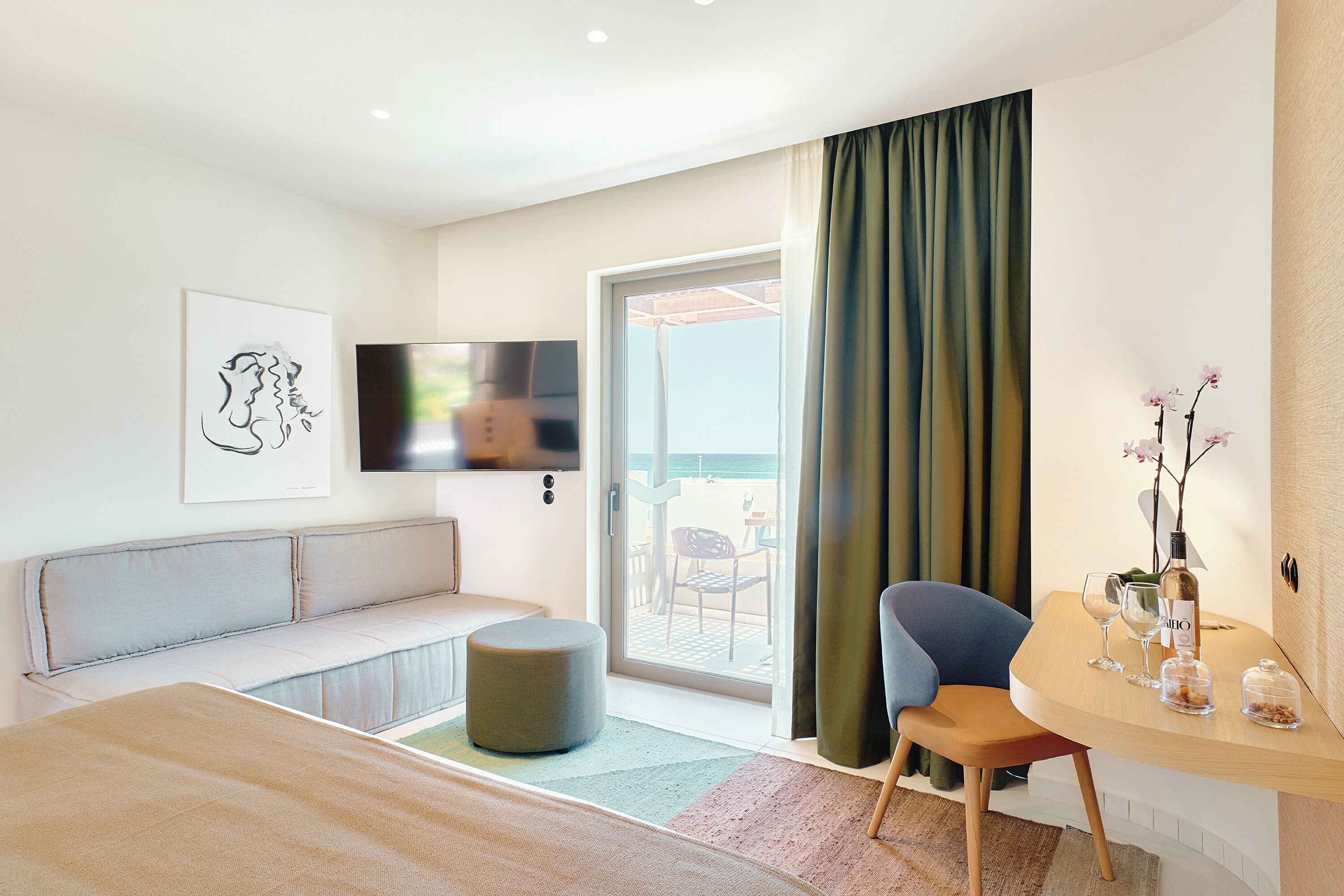 Quarto de hotel moderno com sofá, puf, mesa de jantar e TV fixada na parede.