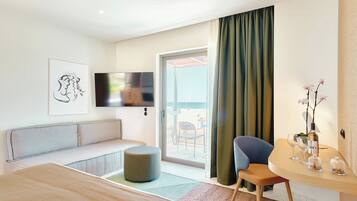 Quarto de hotel moderno com sofá, puf, mesa de jantar e TV fixada na parede.