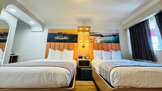 Quarto de hotel com duas camas, um mural da paisagem urbana, cabeceira de madeira, mesa de cabeceira e janela com cortinas.
