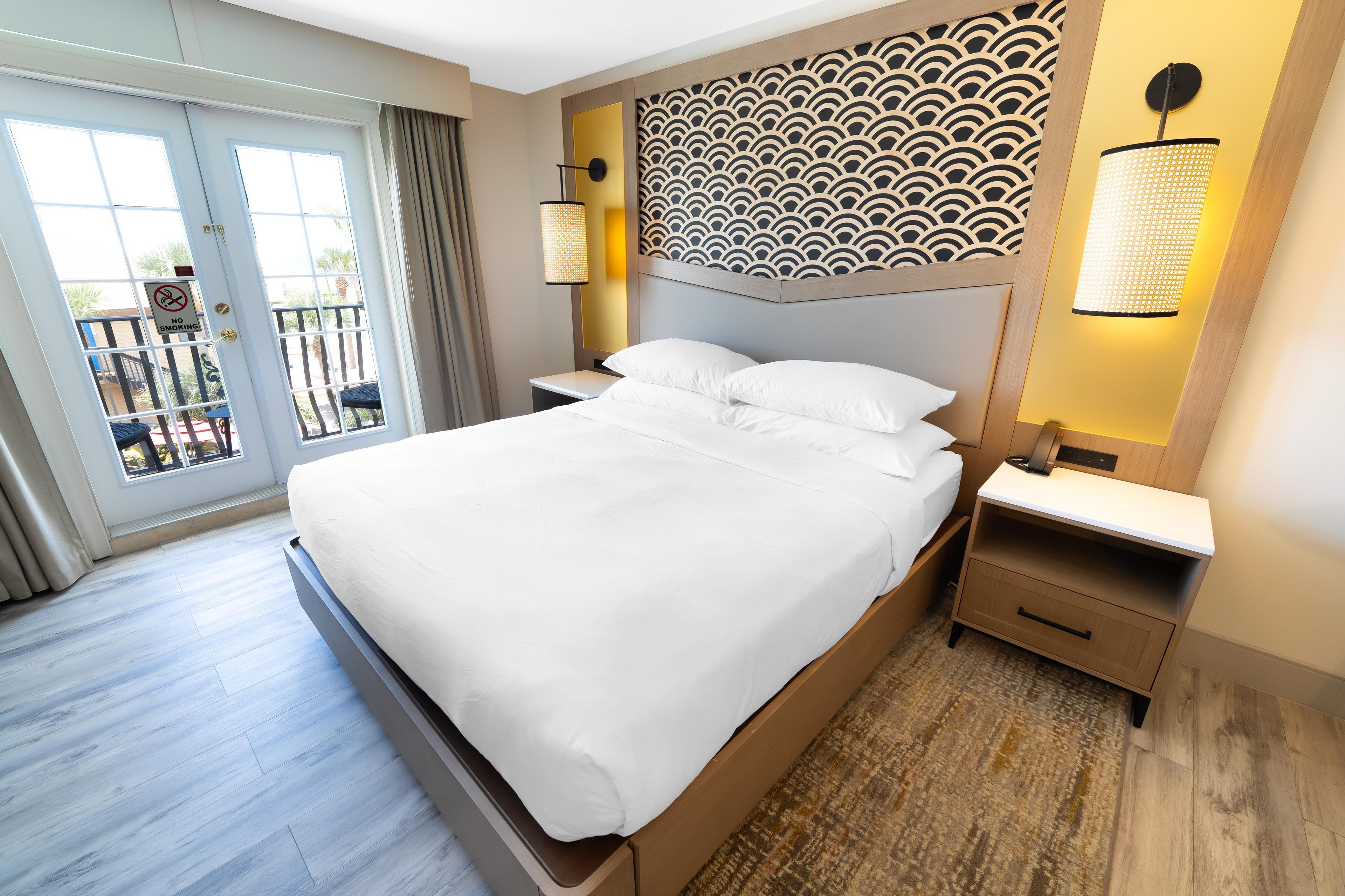 Quarto de hotel com cama, mesas de cabeceira, painel de cabeceira estampado, varanda com vista e placa de 'Proibido Fumar'.