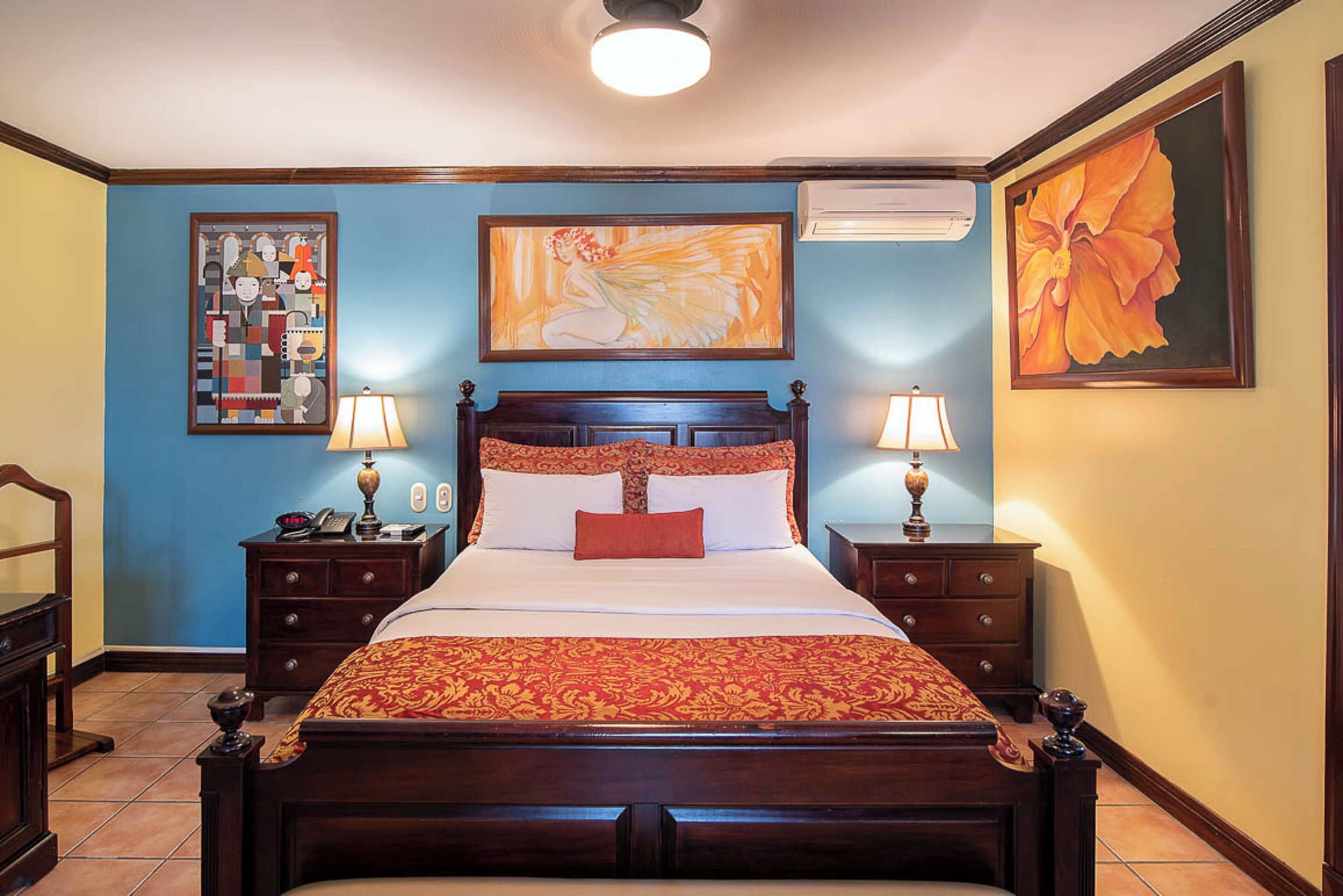 Quarto com uma cama grande, duas mesas de cabeceira e um criado-mudo. Há luminárias na parede e uma luz no teto.