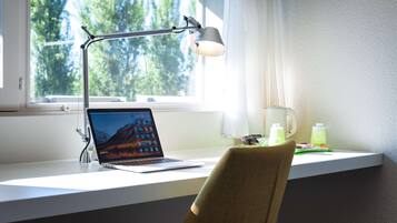 Um laptop numa mesa, próximo a uma janela com vista para árvores, uma luminária e uma caneca.