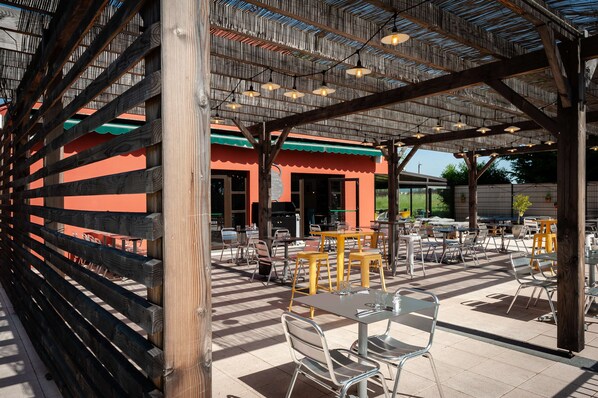 Uma área de pátio externa com mesas e cadeiras, um espaço coberto para sentar e um edifício ao fundo com fachada verde e laranja.