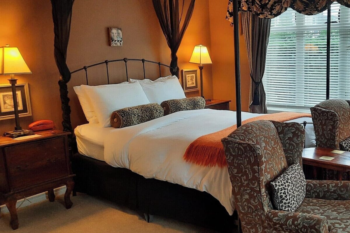 Quarto de hotel com uma cama, duas mesas de cabeceira, um criado-mudo, uma cadeira, uma mesinha e um quadro na parede.