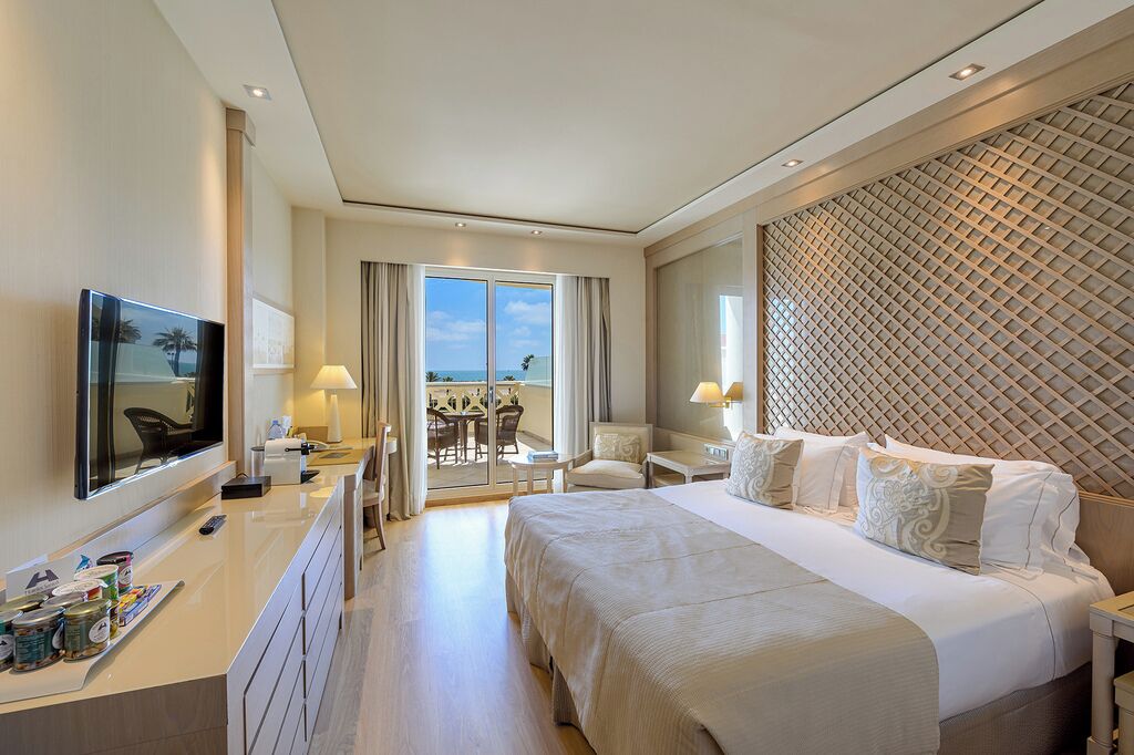 Quarto de hotel com cama, televisão, escrivaninha, varanda com mesa e cadeiras, e vista para o oceano.