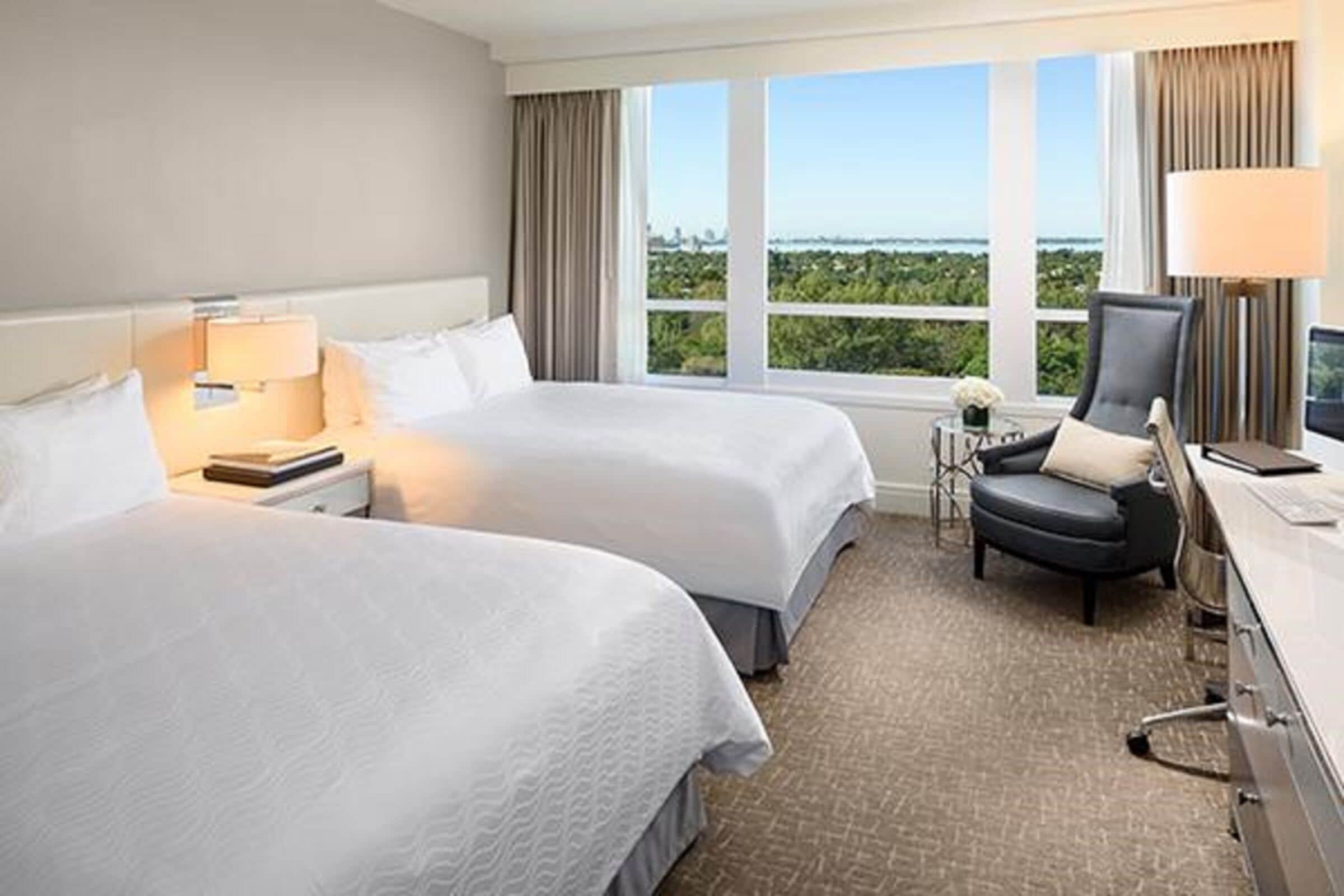 Quarto de hotel com duas camas, uma escrivaninha, uma cadeira, um abajur e vista para o exterior.
