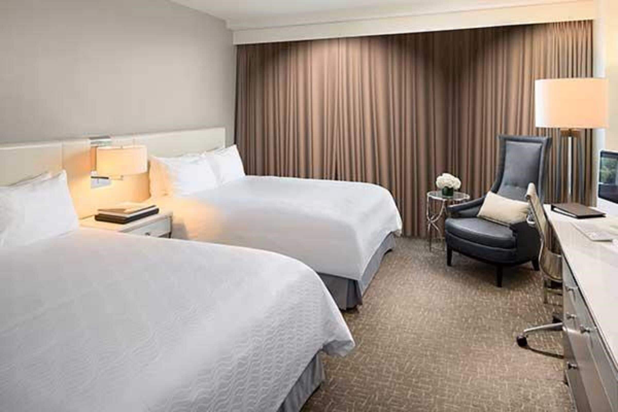Quarto de hotel com duas camas, uma escrivaninha, uma cadeira, um abajur e vista para o exterior.