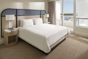 Quarto de hotel com uma cama grande, duas mesas de cabeceira com luminárias, vista para o mar e uma varanda.
