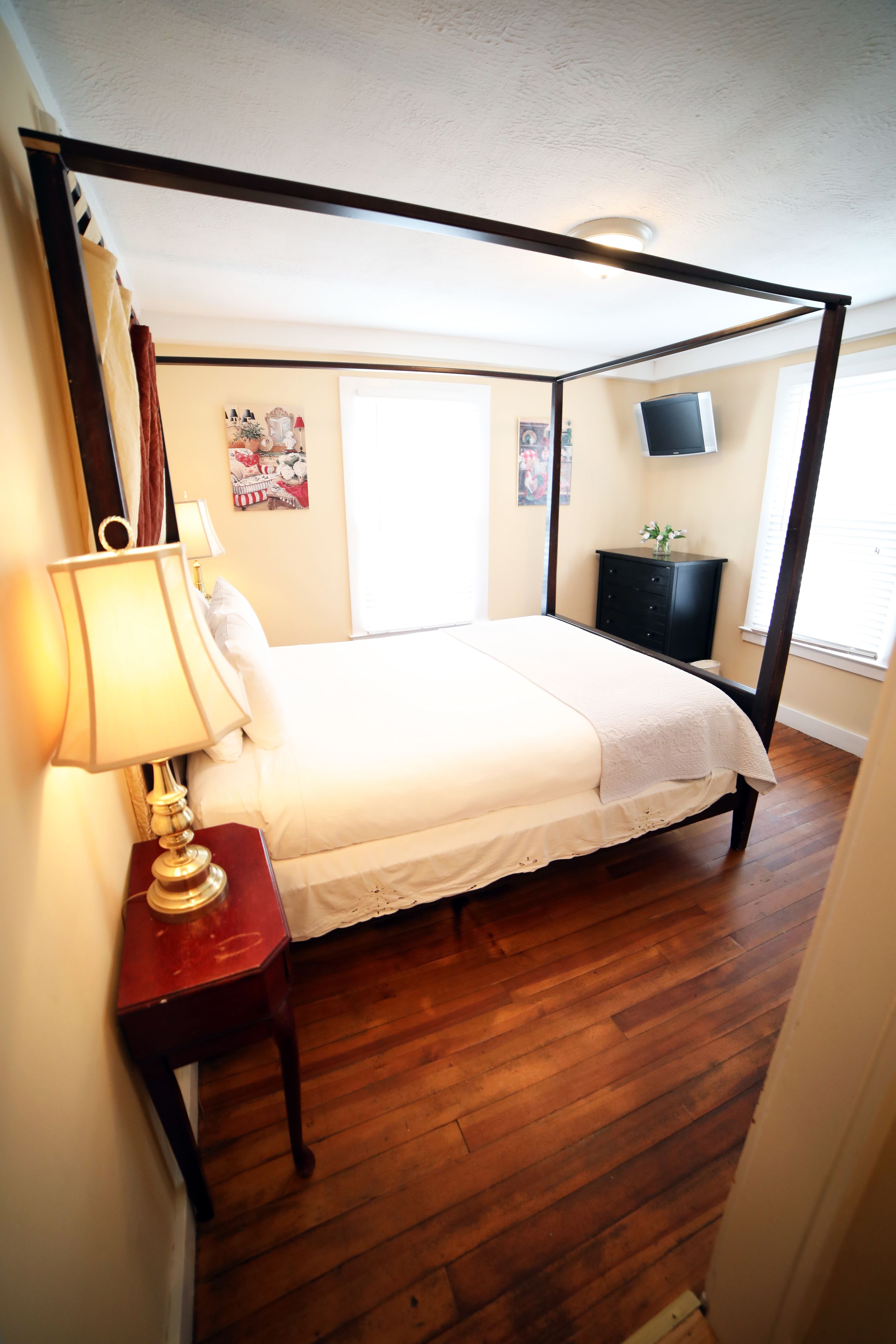 Quarto com cama de dossel, mesa de cabeceira com abajur, cômoda e televisão na parede.