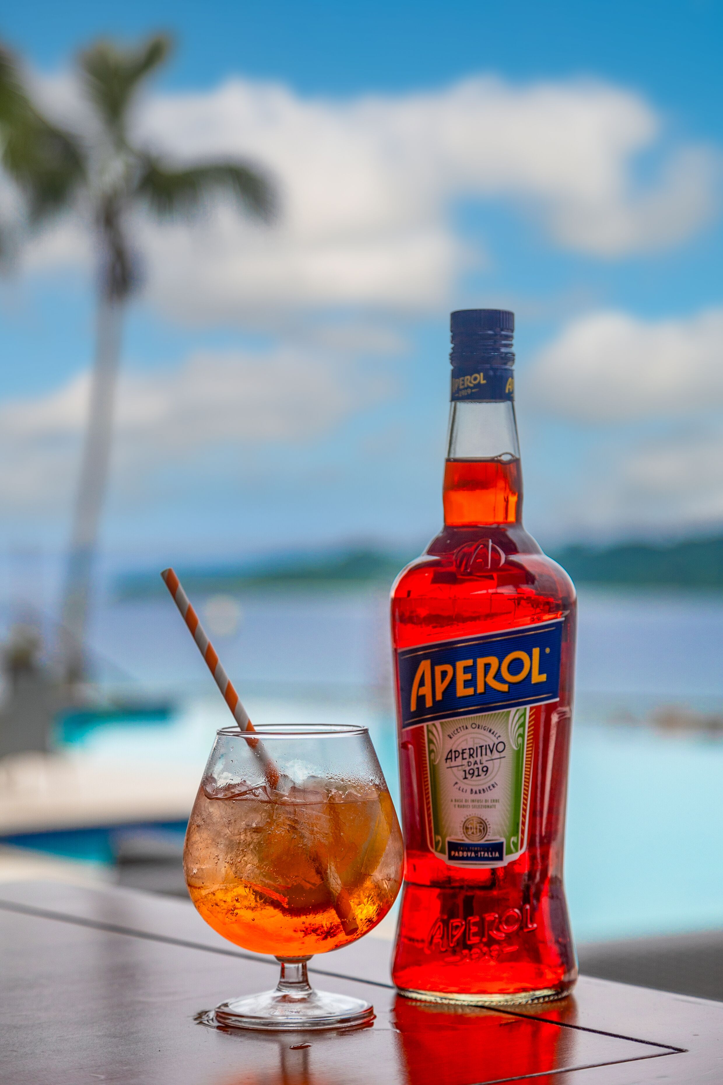 Uma garrafa de Aperol e um copo com canudo sobre uma mesa perto de uma piscina, com uma palmeira ao fundo.