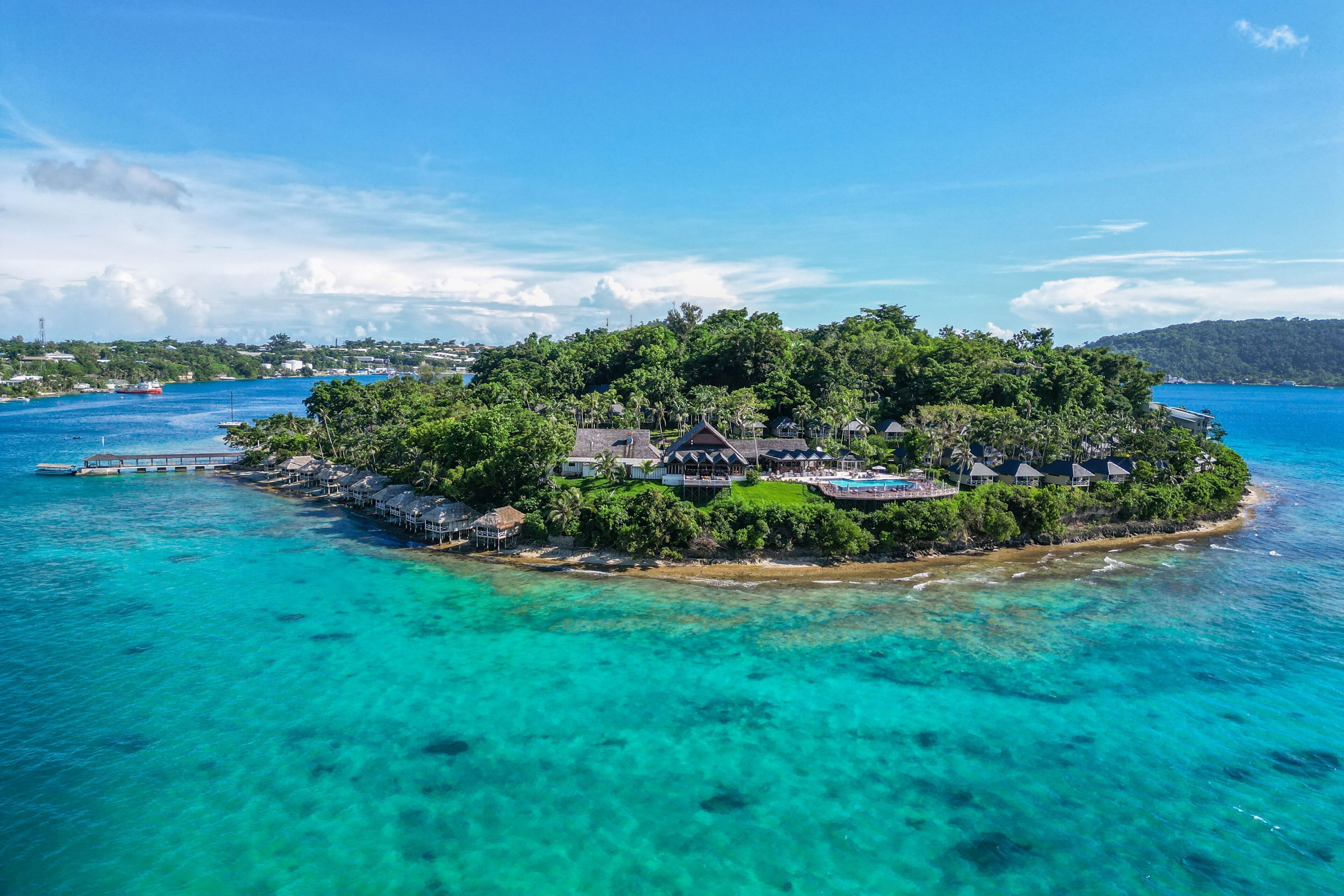 Um resort em ilha com bangalôs sobre a água, piscina e vegetação exuberante.