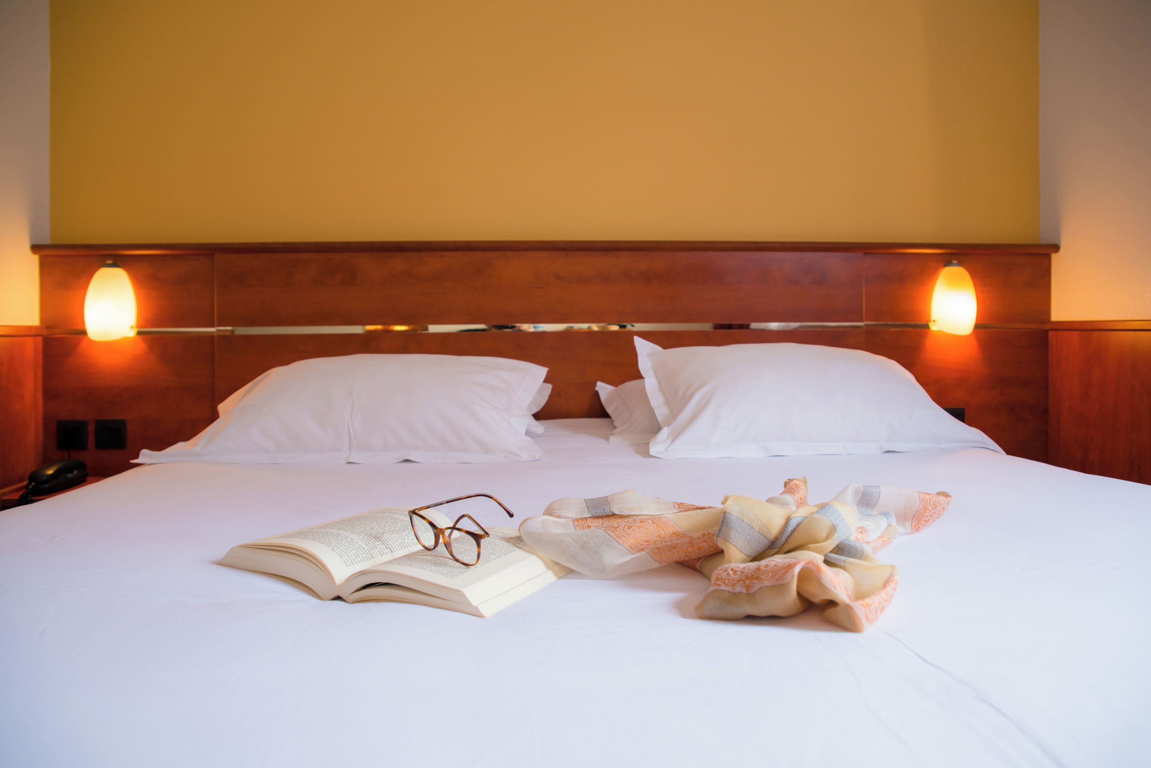 Quarto de hotel com cama, duas luminárias, telefone e um livro.