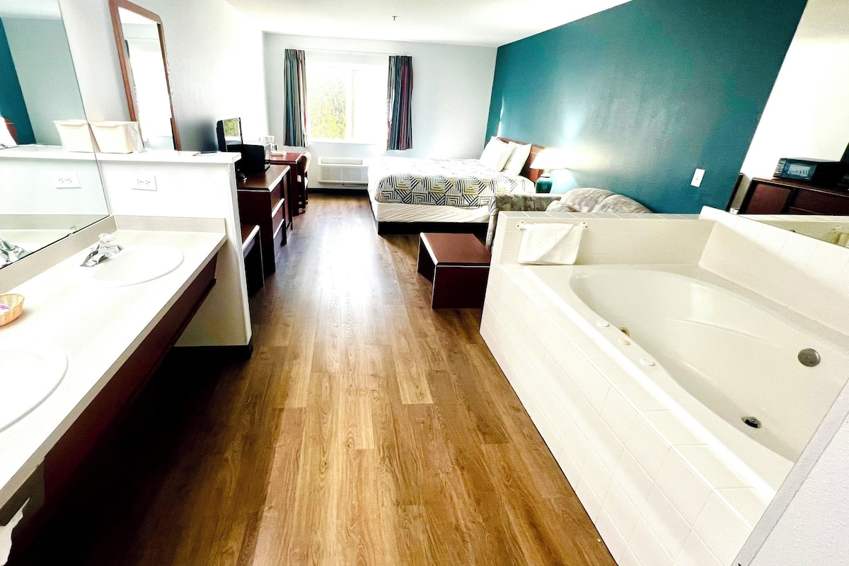 Quarto de hotel com uma banheira grande, um quarto separado com cama e escrivaninha, banheiro com pia e espelho, e janela com cortinas.