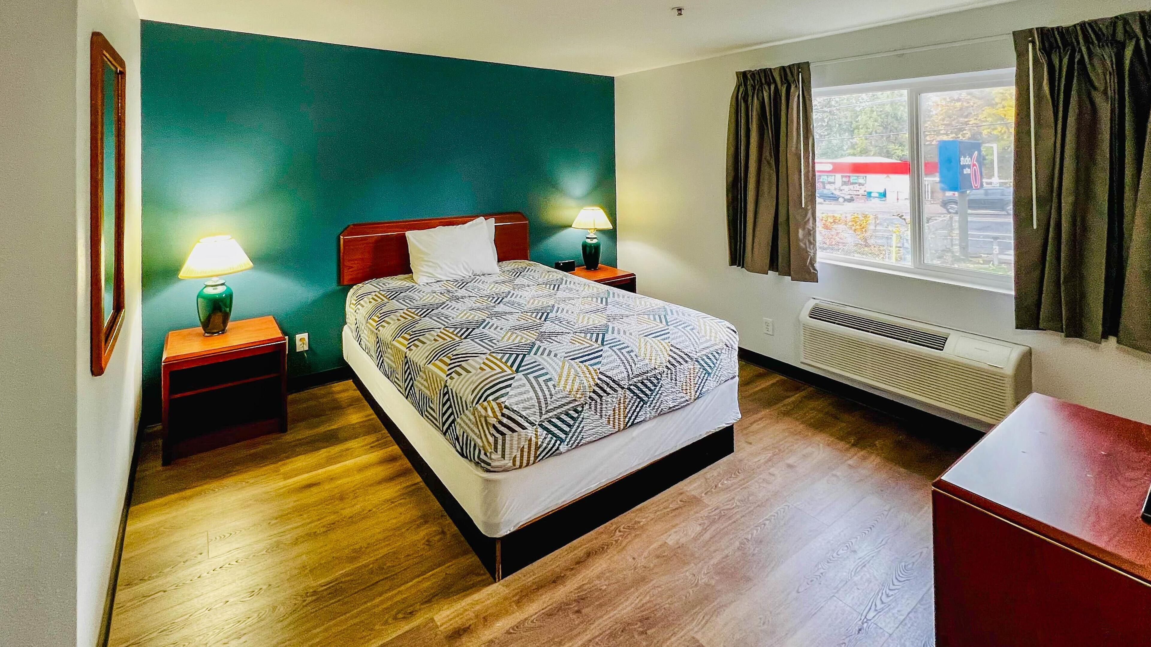 Quarto de hotel com cama, duas mesas de cabeceira com abajures, janela com cortinas e radiador.