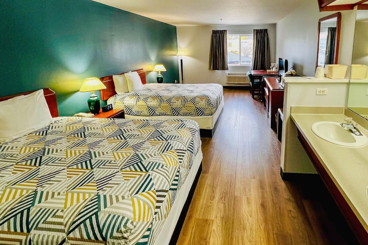 Quarto de hotel com duas camas, uma escrivaninha, um computador, uma luminária de mesa, um espelho e banheiro com pia e espelho.