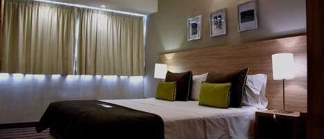 Quarto de hotel com uma cama grande, duas luminárias de cabeceira, uma cabeceira e uma janela com cortinas.