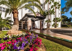 Entrada de hotel com área de espera coberta, palmeiras e canteiro de flores variadas.