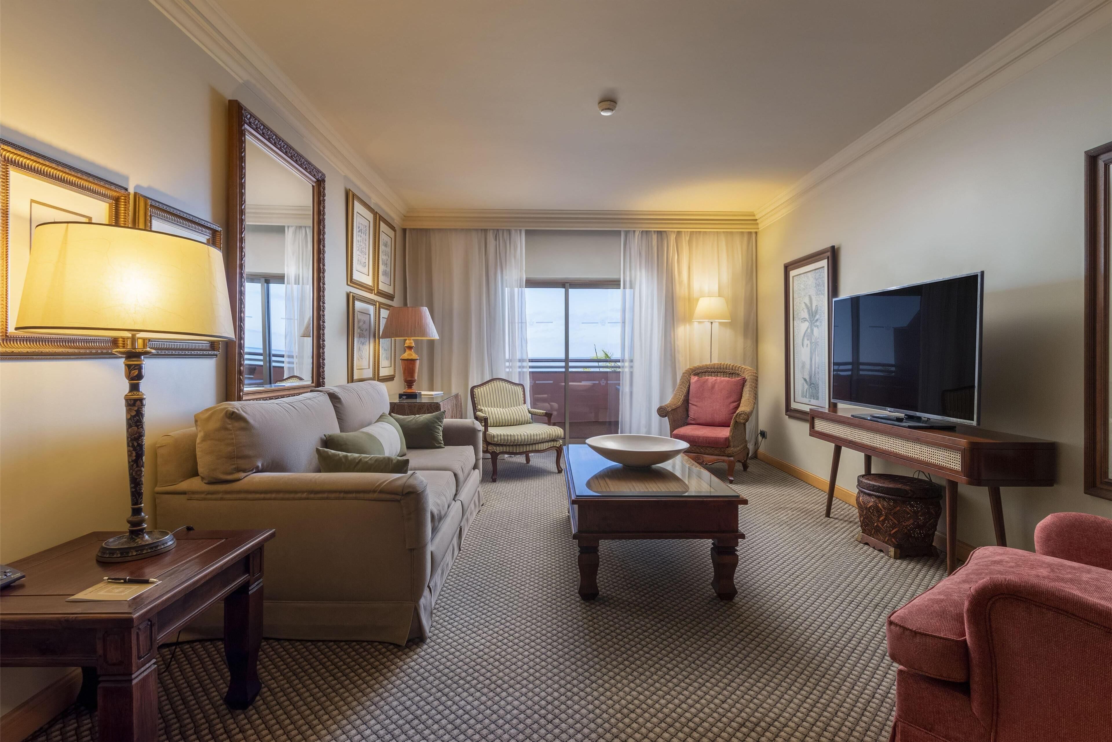 Quarto de hotel com sofá, poltronas, televisão e uma janela grande com cortinas.