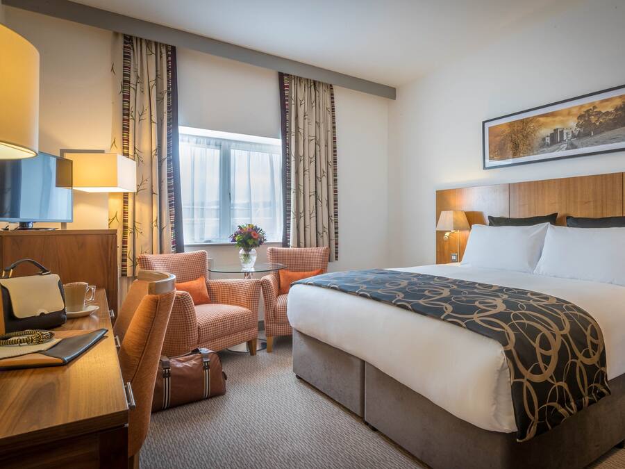 Quarto de hotel com uma cama grande, uma escrivaninha com abajur, uma cadeira, uma mesinha de centro com um vaso e cortinas.