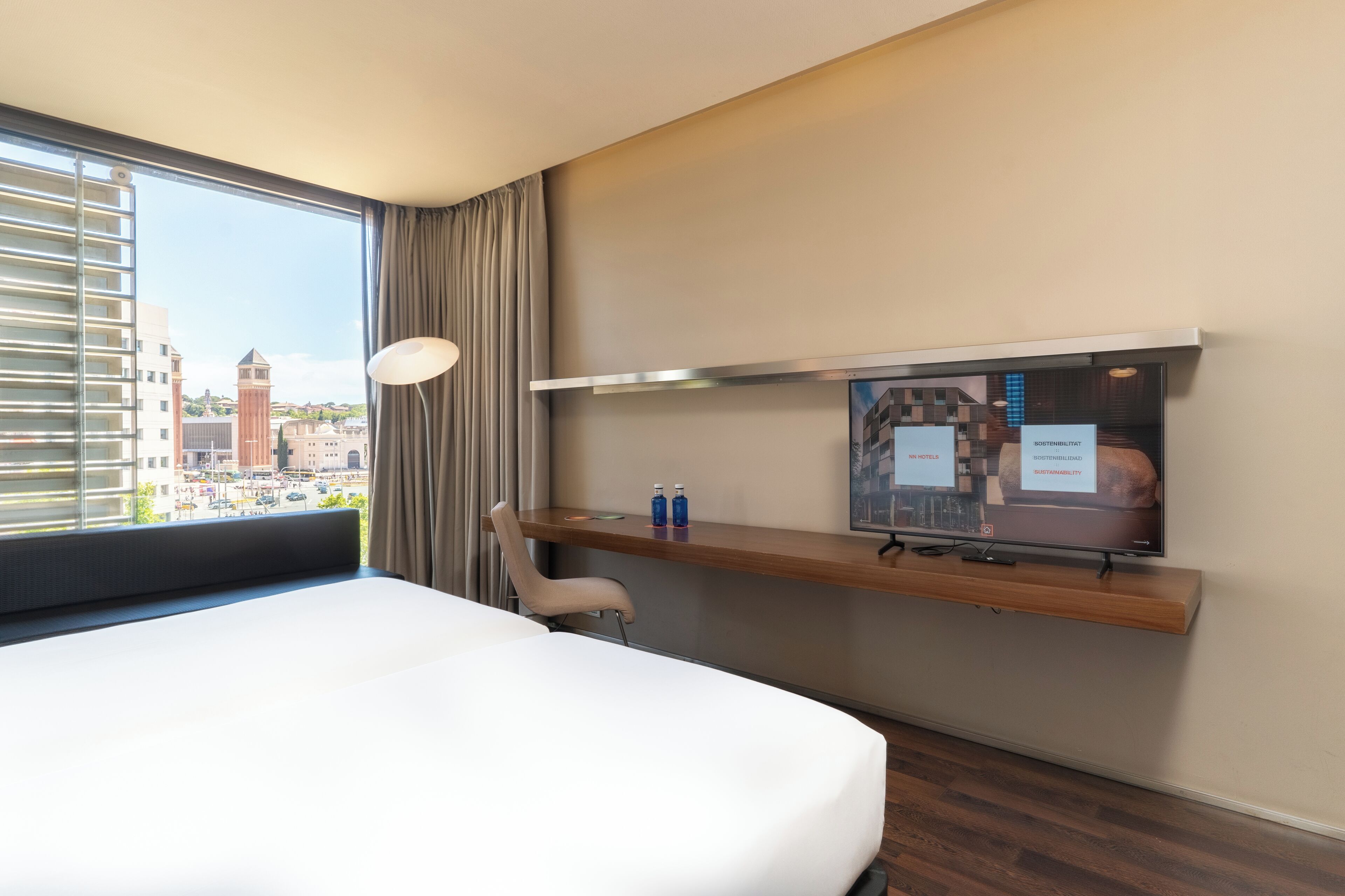 Quarto de hotel com uma cama grande, uma TV em uma prateleira de madeira, uma cadeira e vista para a cidade pela janela.