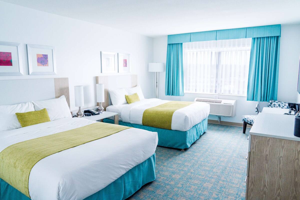 Quarto de hotel com duas camas, uma escrivaninha, uma cadeira, uma mesinha, um abajur e uma janela com cortinas.