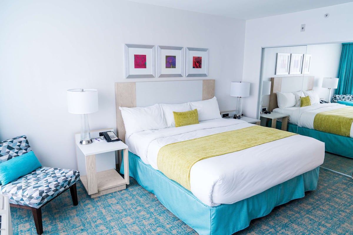 Quarto de hotel com uma cama grande, uma mesinha de cabeceira com abajur, uma cadeira e uma pequena escrivaninha com telefone.