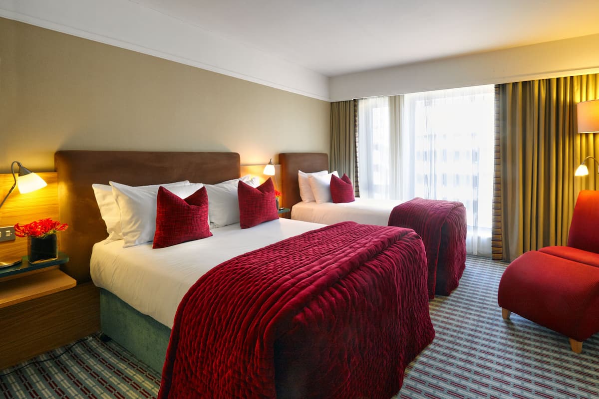 Quarto de hotel com duas camas, uma poltrona vermelha, uma mesa de cabeceira com abajur e vista para a cidade através de grandes janelas.