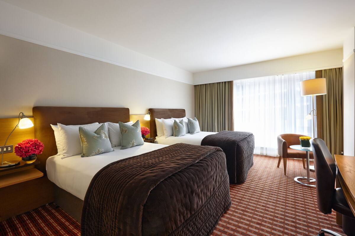 Quarto de hotel com duas camas, uma escrivaninha, uma cadeira e uma luminária.