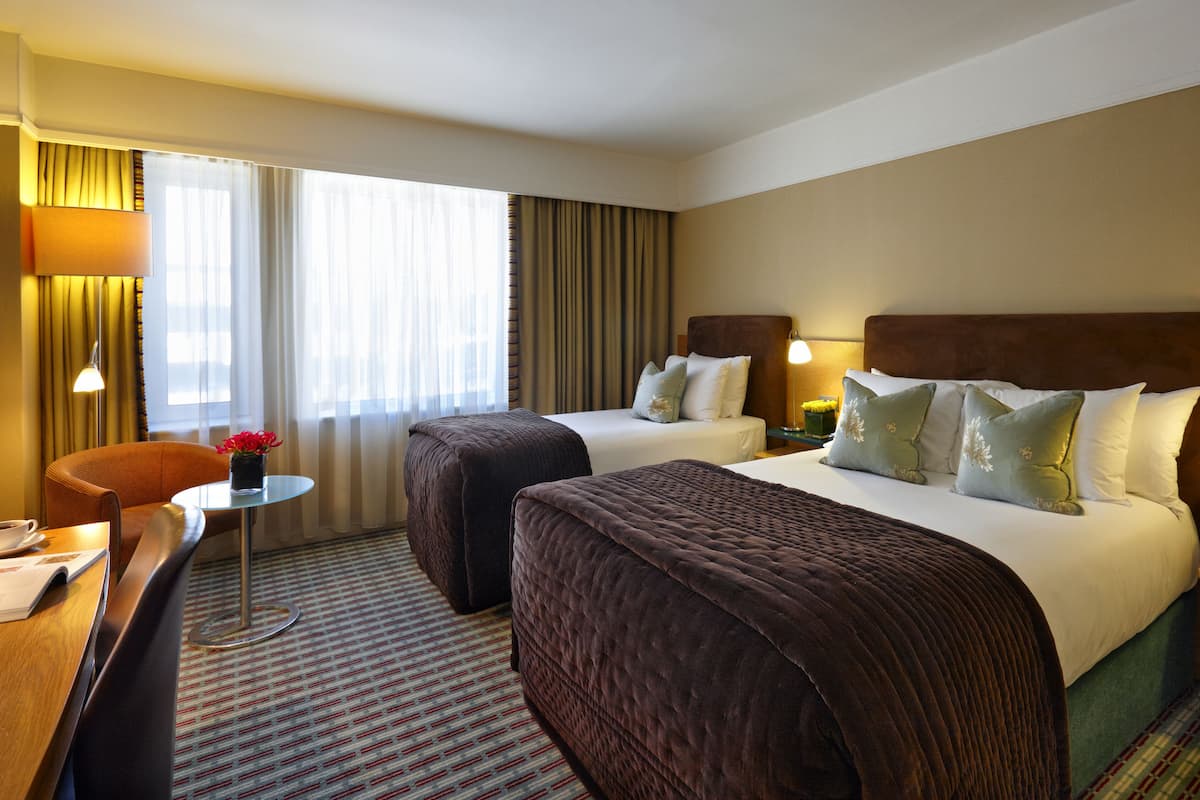 Quarto de hotel com duas camas, uma escrivaninha, uma cadeira e uma janela com cortinas.