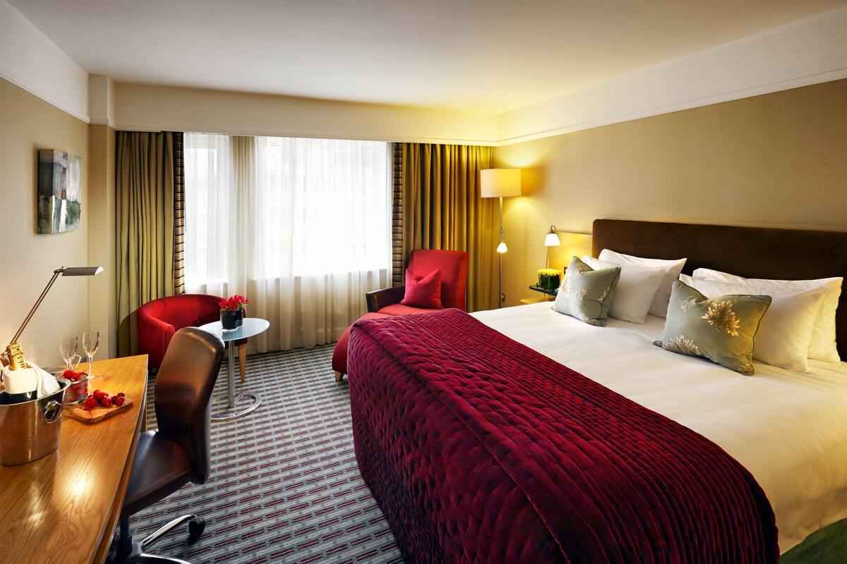 Quarto de hotel com uma cama grande, uma escrivaninha com abajur, uma cadeira, uma mesinha com garrafa e copos, e uma janela com cortinas.